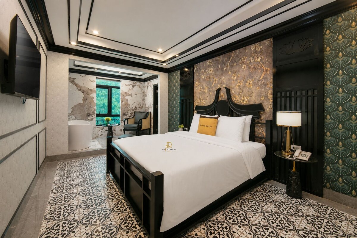 Quarto de hotel com cama grande, mesa de cabeceira com luminária, cadeira e decoração na parede.