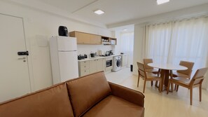 Cozinha moderna com armários brancos, geladeira, micro-ondas e cafeteira. Há uma área de refeições com mesa redonda e cadeiras.