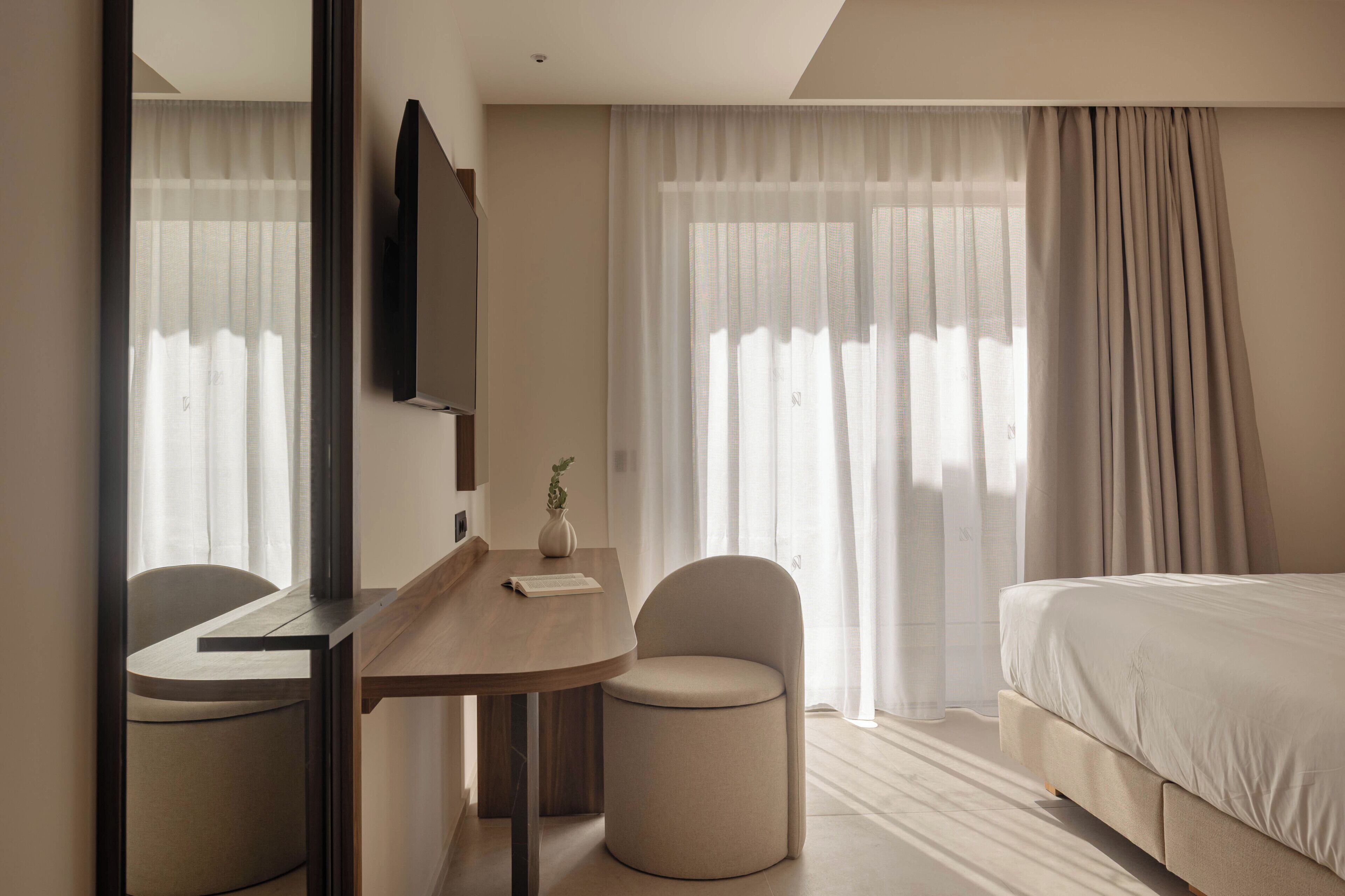 Quarto de hotel com cama, escrivaninha com cadeira, um banquinho, espelho e televisão na parede.