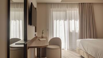 Quarto de hotel com cama, escrivaninha com cadeira, um banquinho, espelho e televisão na parede.