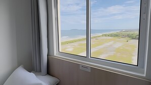 Quarto com uma grande janela com vista para uma praia e montanhas ao longe.