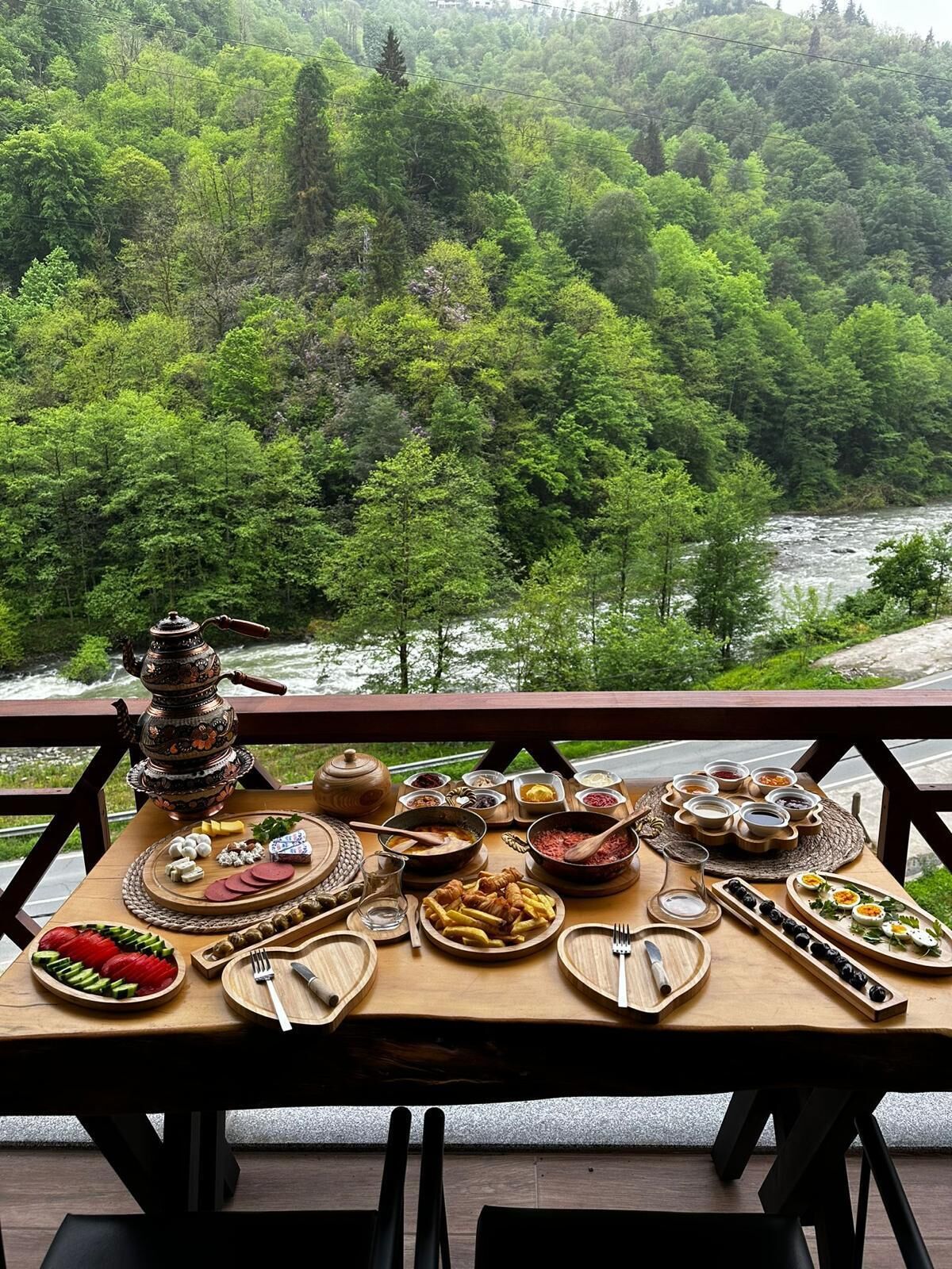 Mesa posta com diversos alimentos e um bule, com vista para uma área arborizada e um rio.