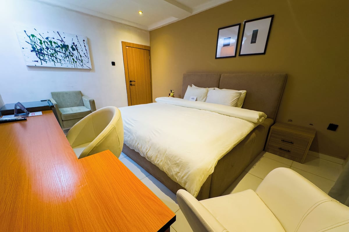 Quarto de hotel com cama, escrivaninha, cadeira, mesa de cabeceira e porta.