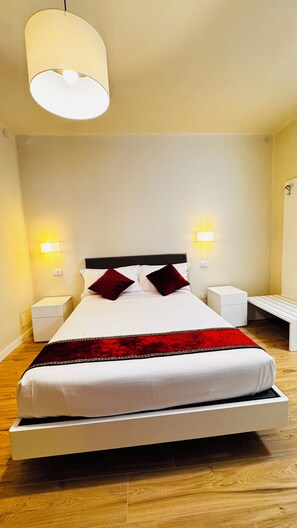 Cama bem arrumada com colcha branca e vermelha, duas mesas de cabeceira com luminárias e um lustre.