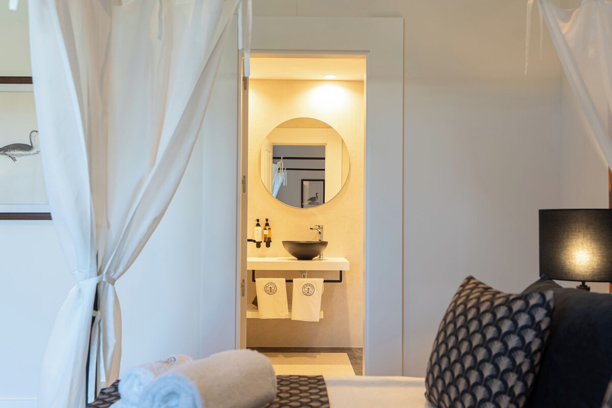 Quarto de hotel com uma cama, mesa de cabeceira, luminária, banheiro com pia e espelho, e cortina.