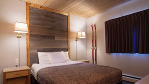 Quarto de hotel com cabeceira de madeira, duas luminárias de mesa, cama com colcha estampada em tons de castanho e uma janela com cortinas.