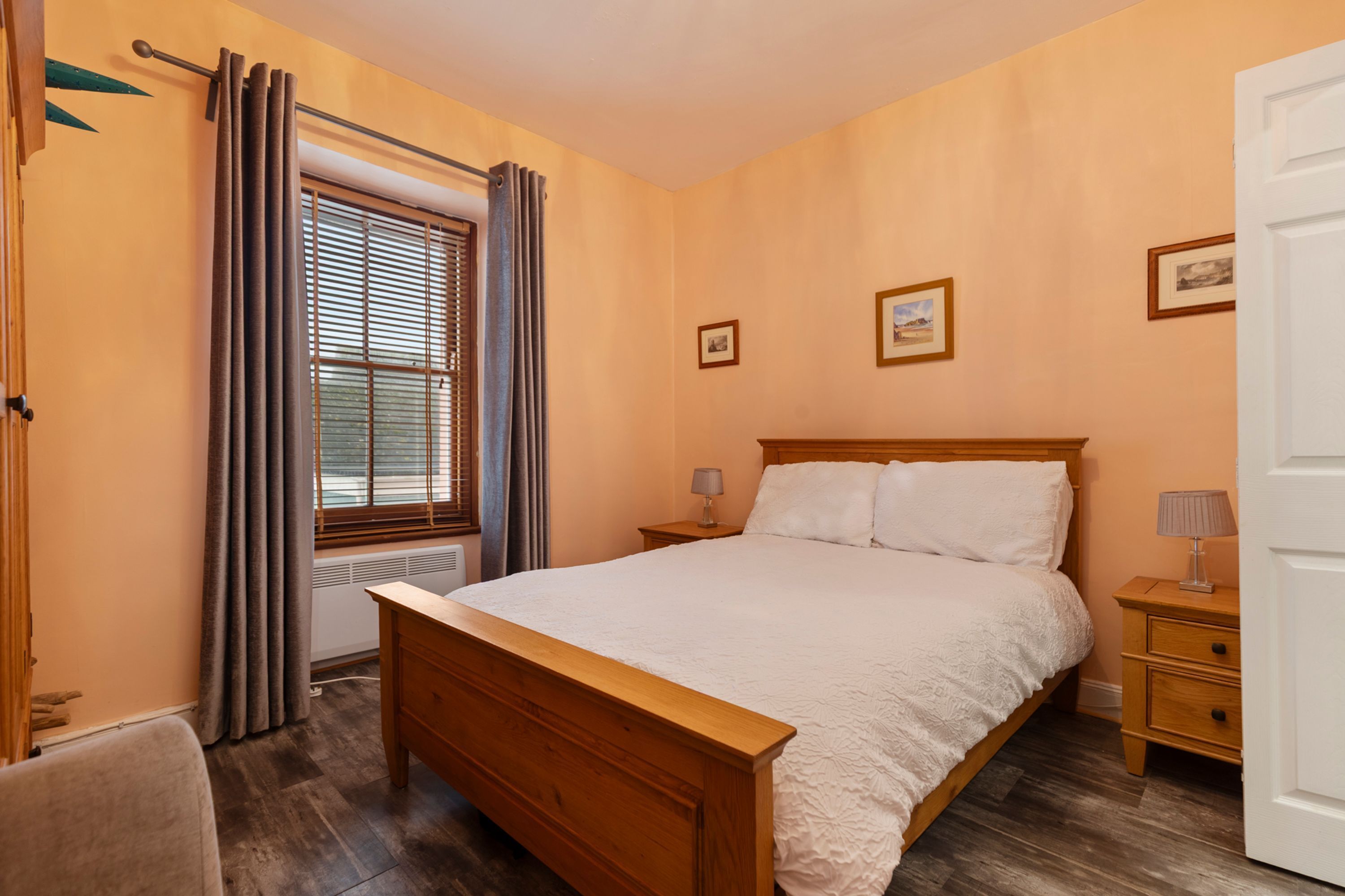 Quarto com cama de madeira, mesa de cabeceira com abajur, janela com persianas e porta.