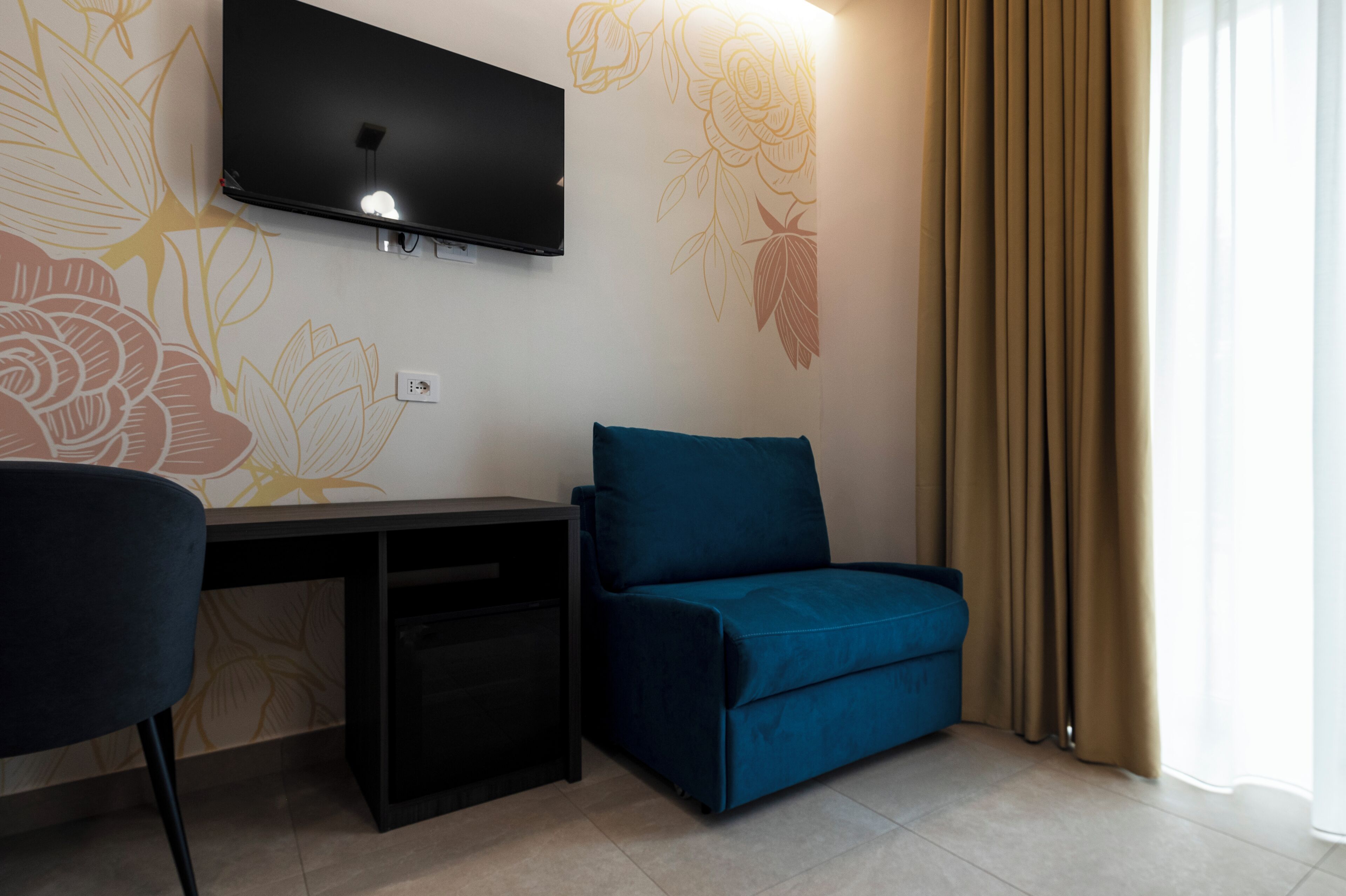 Quarto com televisão de tela plana na parede, sofá azul, escrivaninha com cadeira e janela com cortinas.