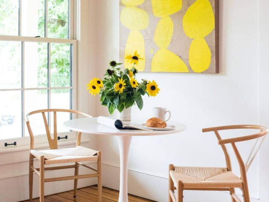 Um quarto com uma mesa redonda branca, duas cadeiras de madeira, um vaso com girassóis e um quadro abstrato amarelo na parede.