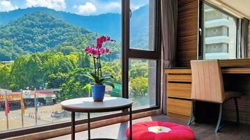 Quarto com uma janela grande, uma mesinha com uma planta, um almofado vermelho e uma escrivaninha de madeira com uma cadeira.