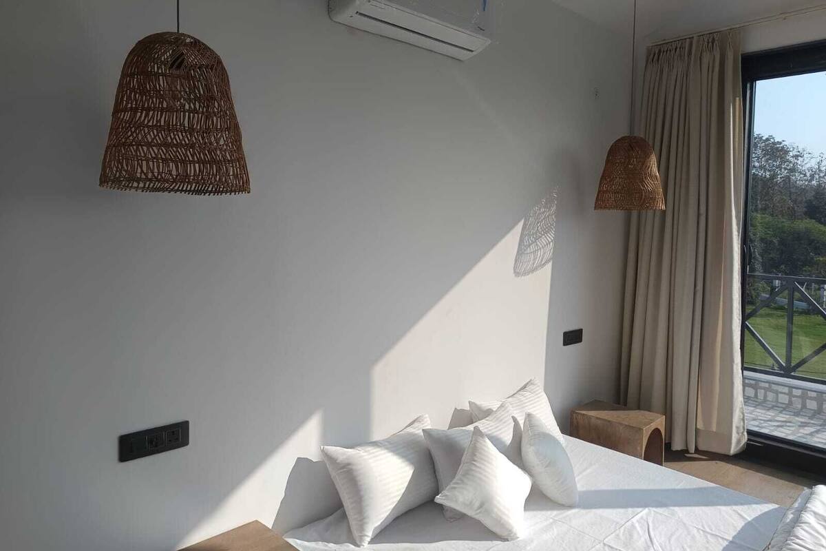 Quarto com cama, almofadas brancas, um banquinho de madeira e duas luminárias pendentes feitas de tecido.