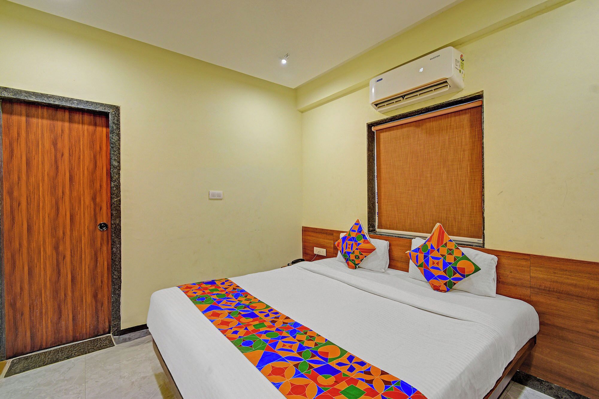 Quarto de hotel com cama, porta de madeira, aparelho de ar condicionado e janela com persiana de madeira.
