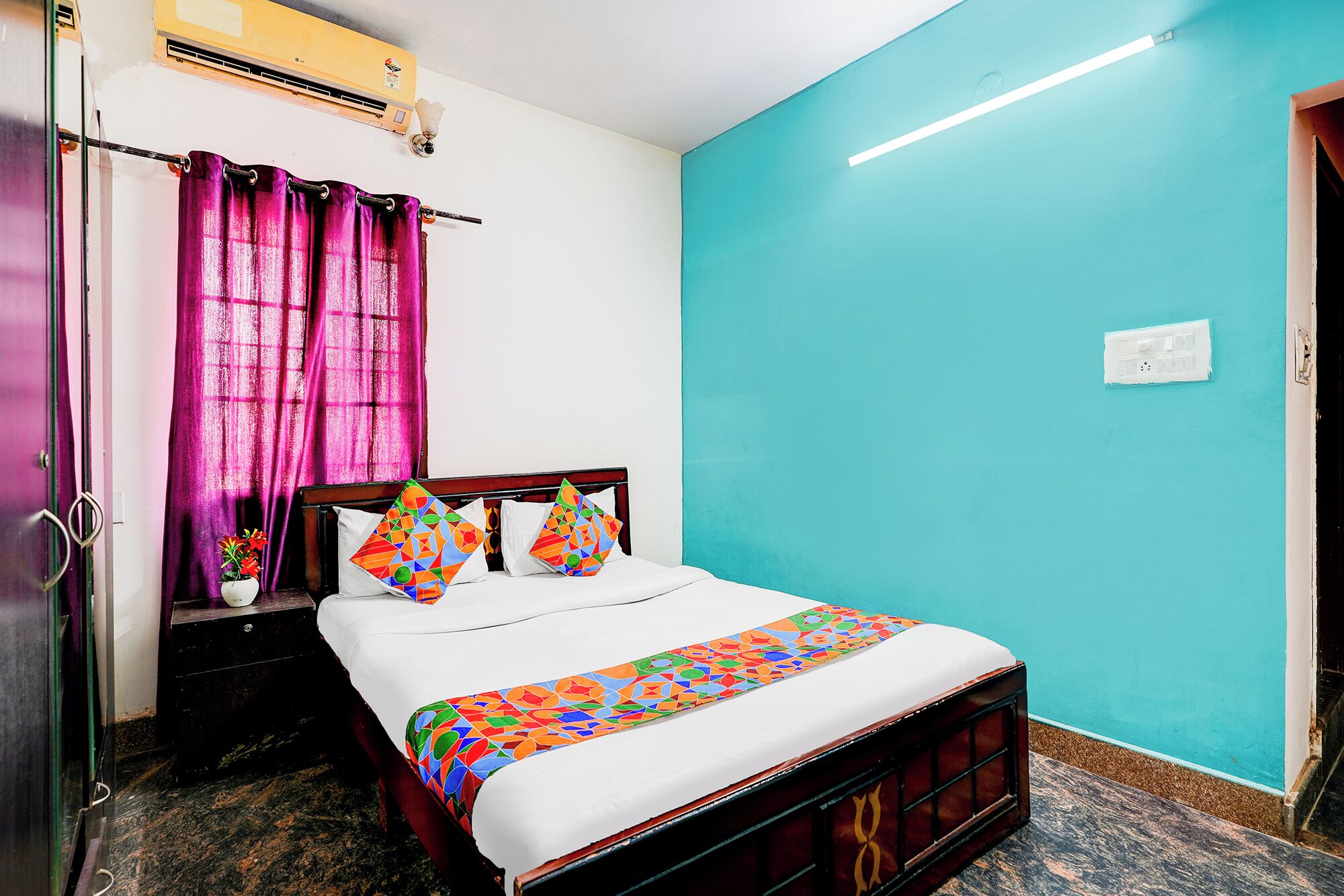 Quarto com cama, mesa de cabeceira, ar condicionado na parede, porta e janela com cortinas.