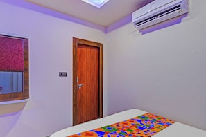 Quarto com porta de madeira, ar condicionado, cama com colcha de padrão geométrico colorido e luminária pequena fixada na parede.