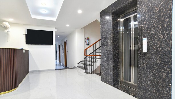 Um lobby de hotel moderno com um balcão de recepção de mármore, uma escadaria com elevador de vidro e um extintor de incêndio fixado na parede.
