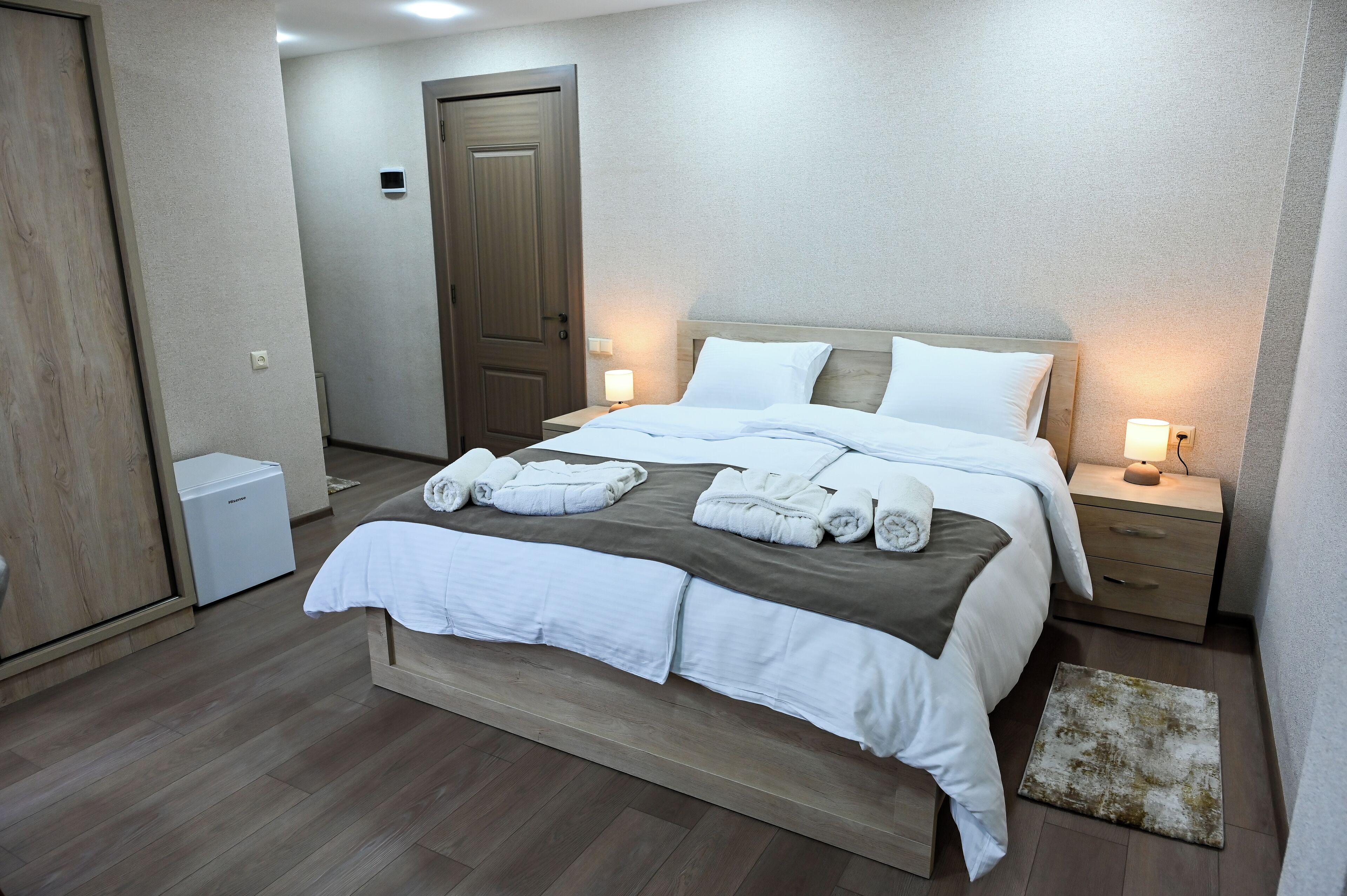 Uma cama bem arrumada com lençóis e toalhas brancas, cabeceira de madeira, mesa de cabeceira com abajur e um pequeno cesto de lixo branco.