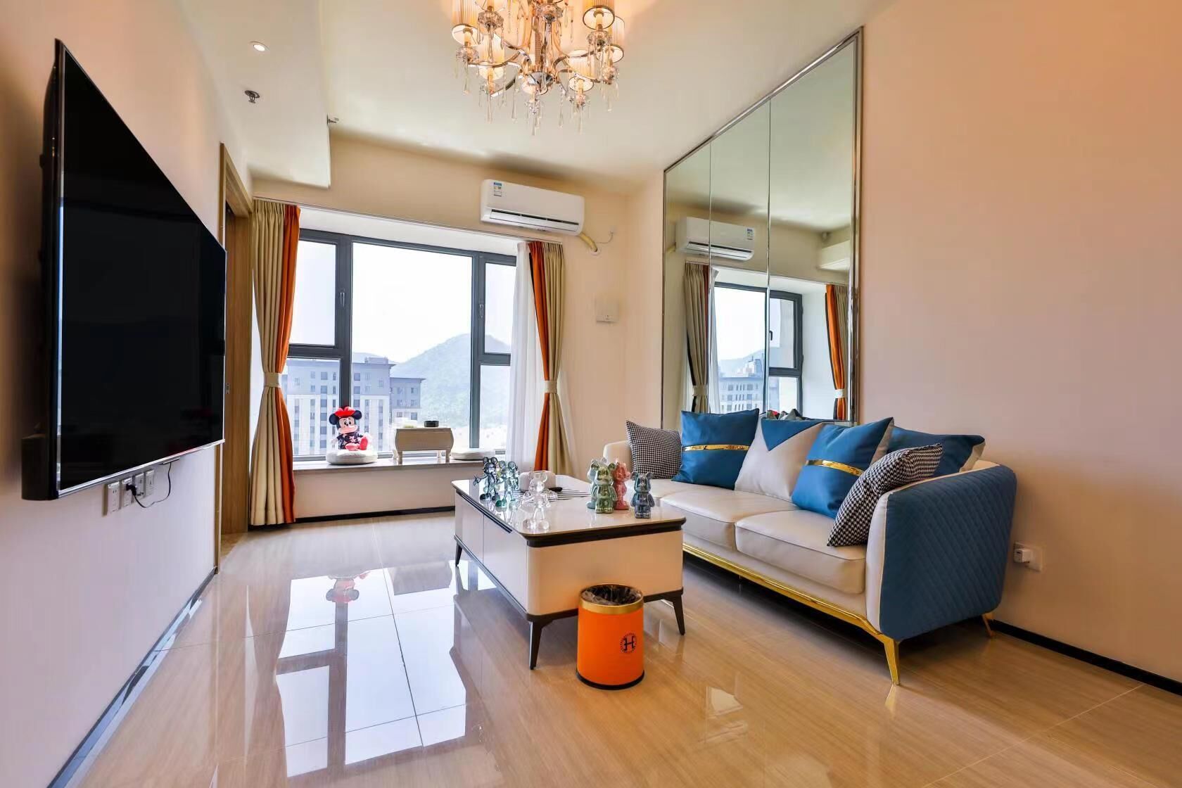 Uma sala de estar moderna com uma TV de tela plana, um sofá com almofadas azuis, uma mesa de centro com objetos decorativos e grandes janelas com cortinas.