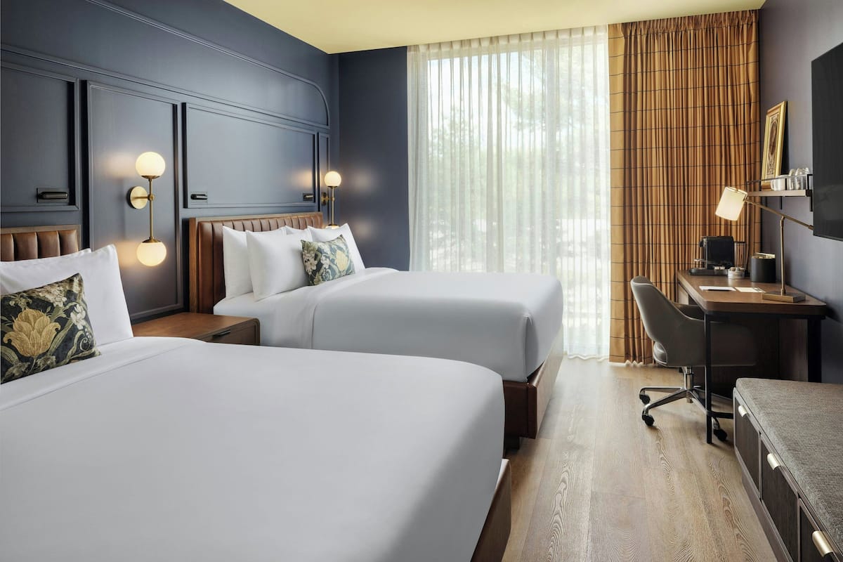 Quarto de hotel com duas camas, uma escrivaninha, uma cadeira, uma televisão e uma janela com cortinas.