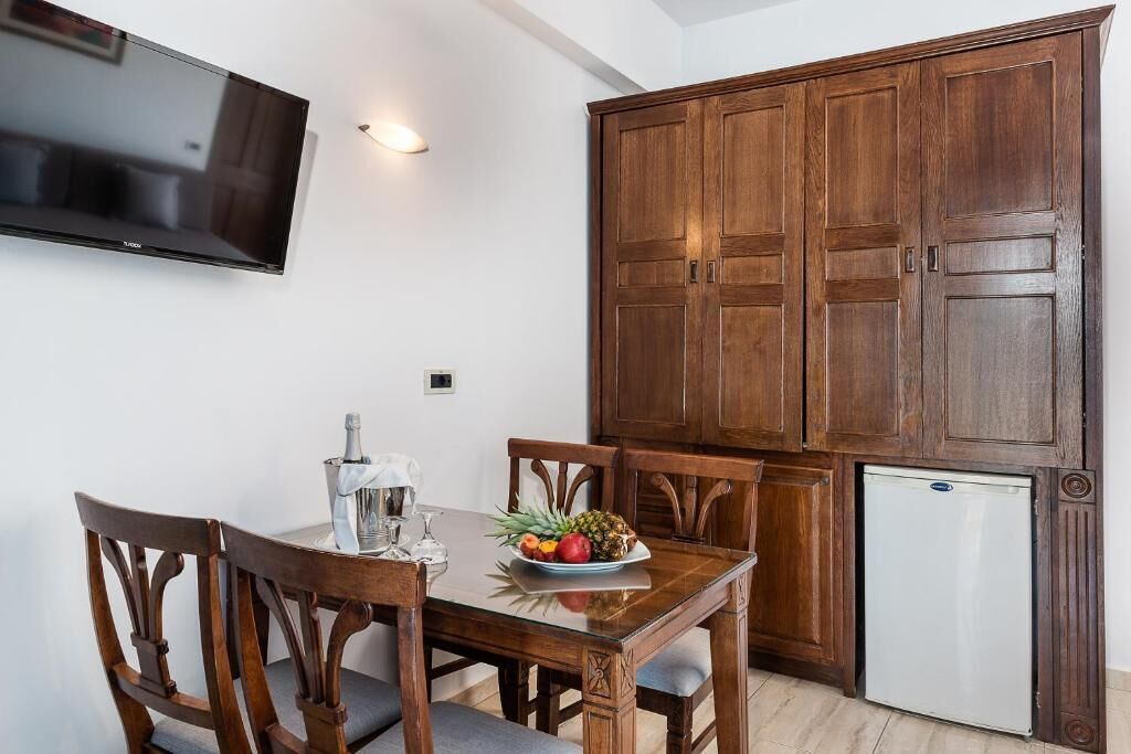 Uma sala de jantar com mesa de madeira, cadeiras, um pequeno frigorífico e uma televisão na parede.