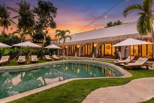 Uma área de piscina com espreguiçadeiras e guarda-sóis, rodeada por palmeiras e um edifício com telhado de palha.