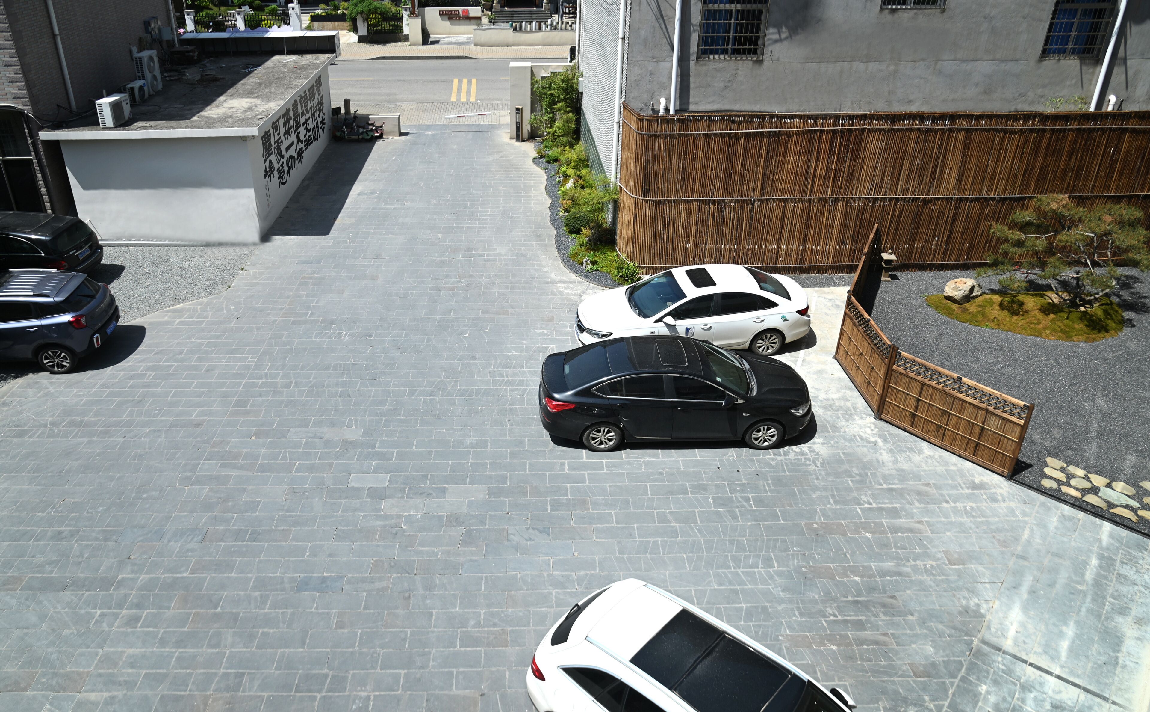 Uma área de estacionamento com dois carros, um edifício de fachada cinzenta e uma cerca de bambu.