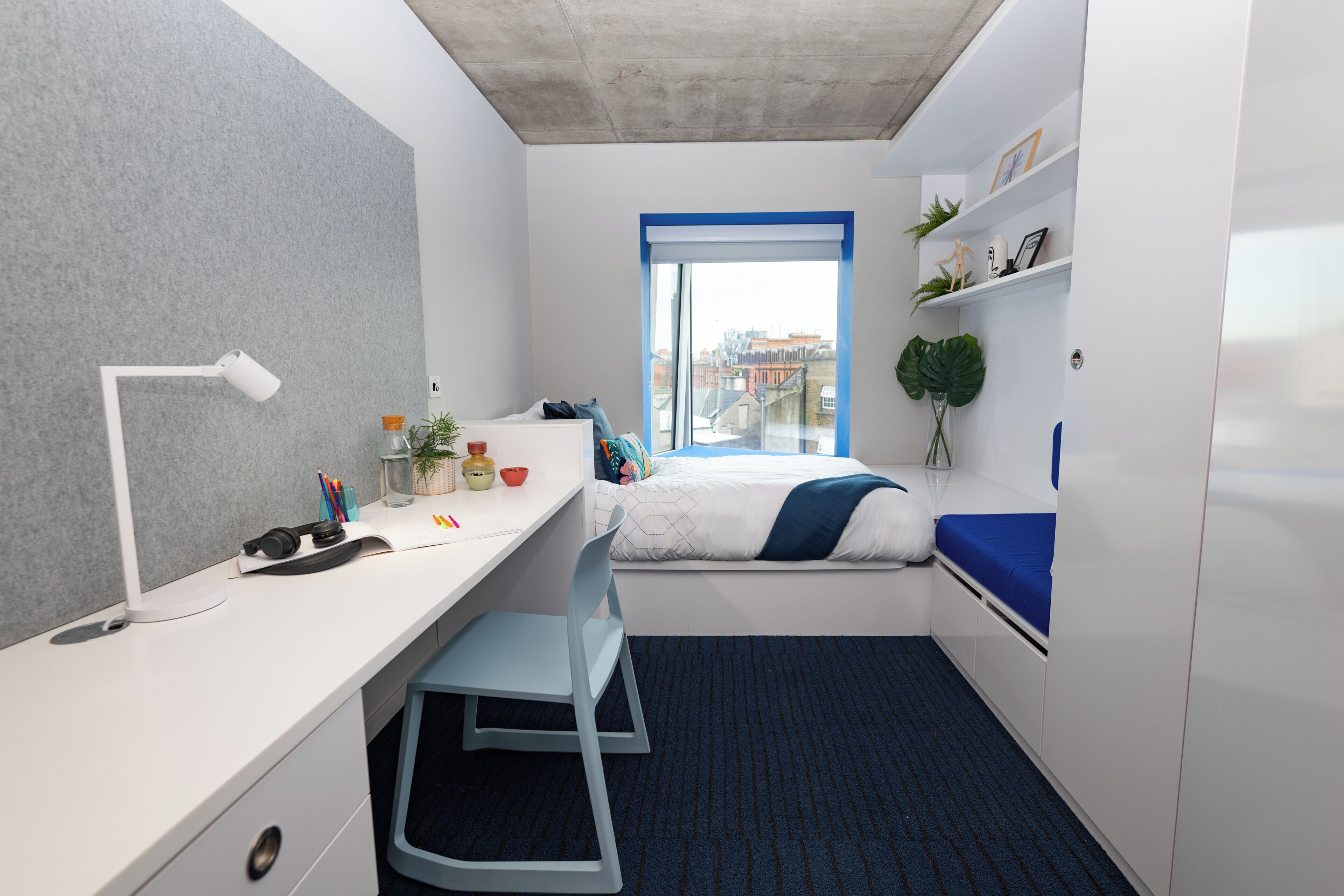Quarto compacto com escrivaninha, cadeira, cama e janela com vista para edifícios.