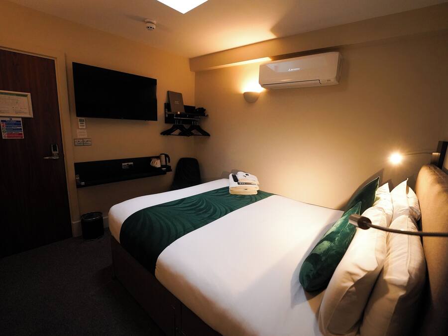 Quarto de hotel com cama, televisão na parede, ar condicionado, escrivaninha com um laptop e uma lixeira.
