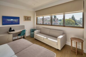 Quarto de hotel moderno com cama, sofá, escrivaninha com cadeira, televisão e uma grande janela com vista para a cidade.