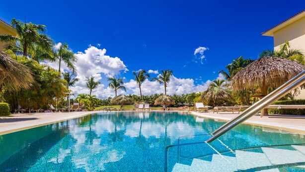 Piscina azul cristalina com escada de metal, cercada por palmeiras e cadeiras de descanso, sob um céu azul vibrante.