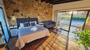 Quarto com parede de pedra, uma cama com almofadas, um sofá, uma mesa pequena e vista para o verde através de uma porta de vidro.