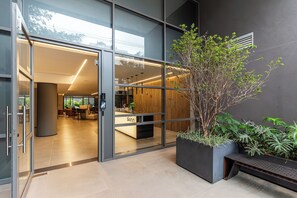 Entrada de um edifício moderno com portas de vidro, balcão de recepção e plantas em vasos.