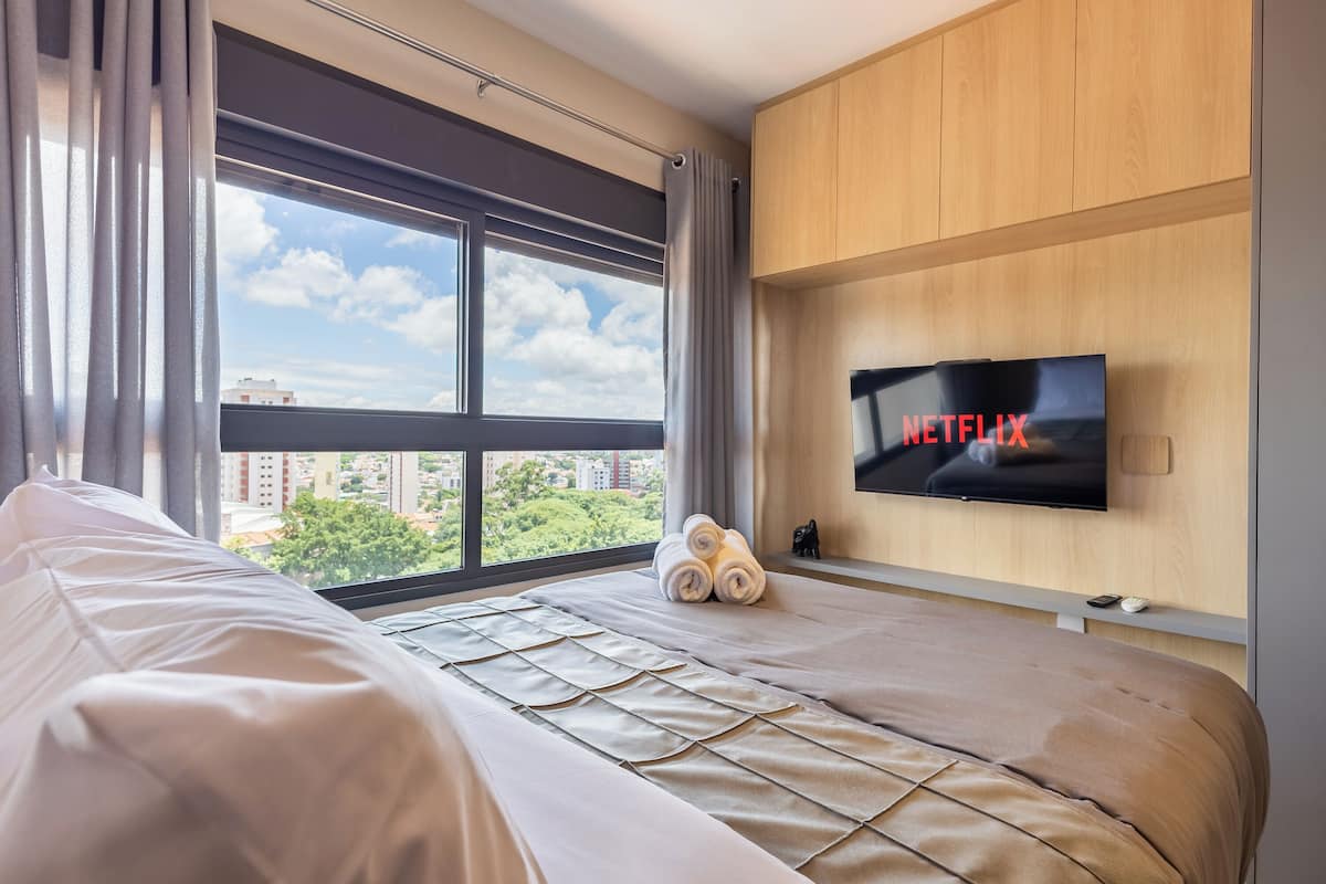 Quarto de hotel moderno com uma grande janela, uma cama com colcha bege, uma TV de tela plana na parede e vista para a paisagem urbana.