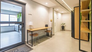 Um saguão moderno de hotel com um balcão de recepção, duas cadeiras, uma pequena planta e uma área de lavanderia visível.