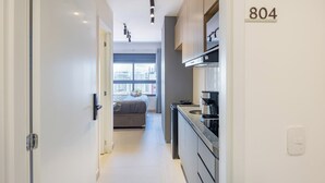 Quarto de hotel moderno com cozinha compacta, cama com vista e paisagem urbana visível pela janela.