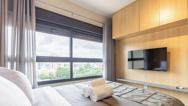 Quarto de hotel com uma janela grande, uma televisão de tela plana na parede e toalhas dobradas na cama.
