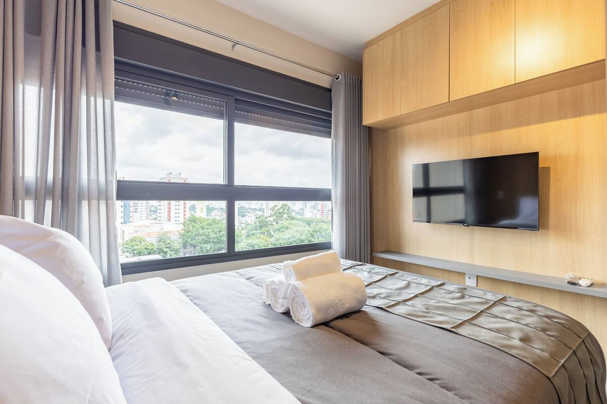 Quarto de hotel com uma janela grande, uma televisão de tela plana na parede e toalhas dobradas na cama.