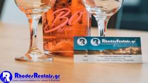 Uma garrafa de vinho B&O Heme ao lado de dois copos sobre uma mesa, com um folheto e o logotipo da RhodesRentals.gr visíveis.