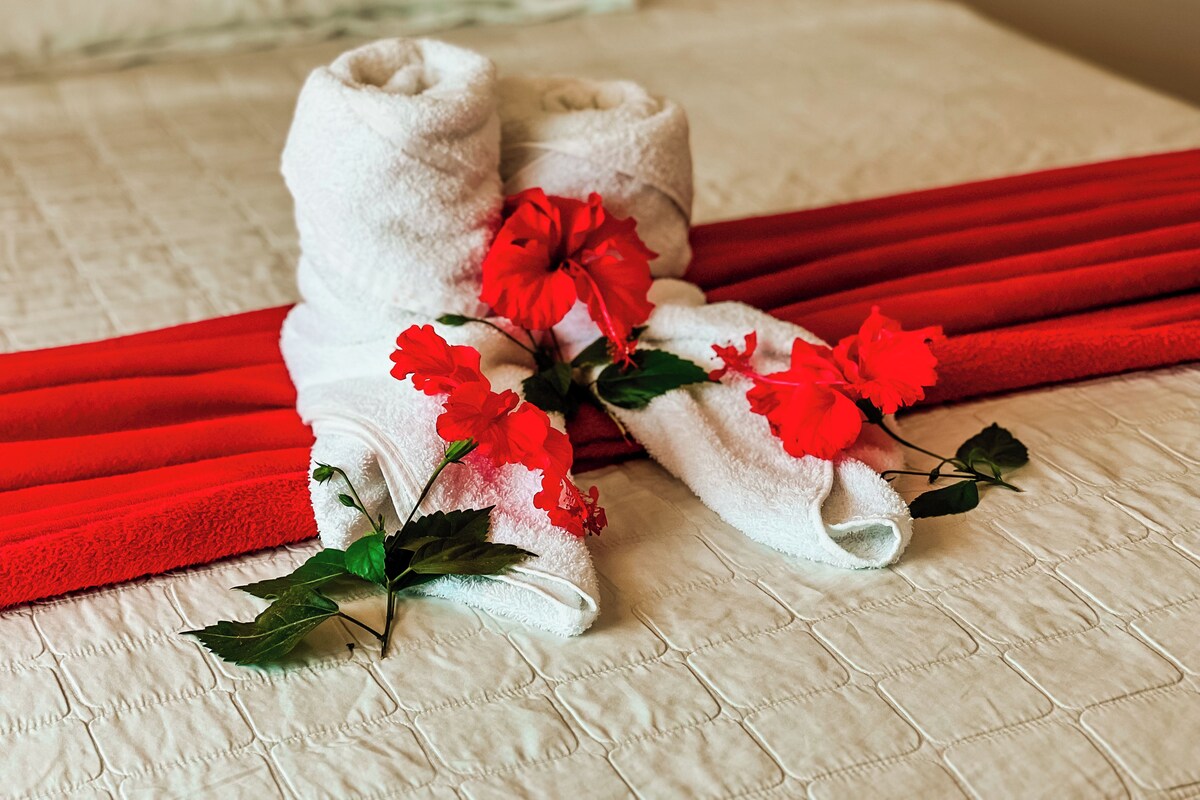 Uma cama bem arrumada com edredom branco, almofadas decorativas vermelhas e um laço vermelho.