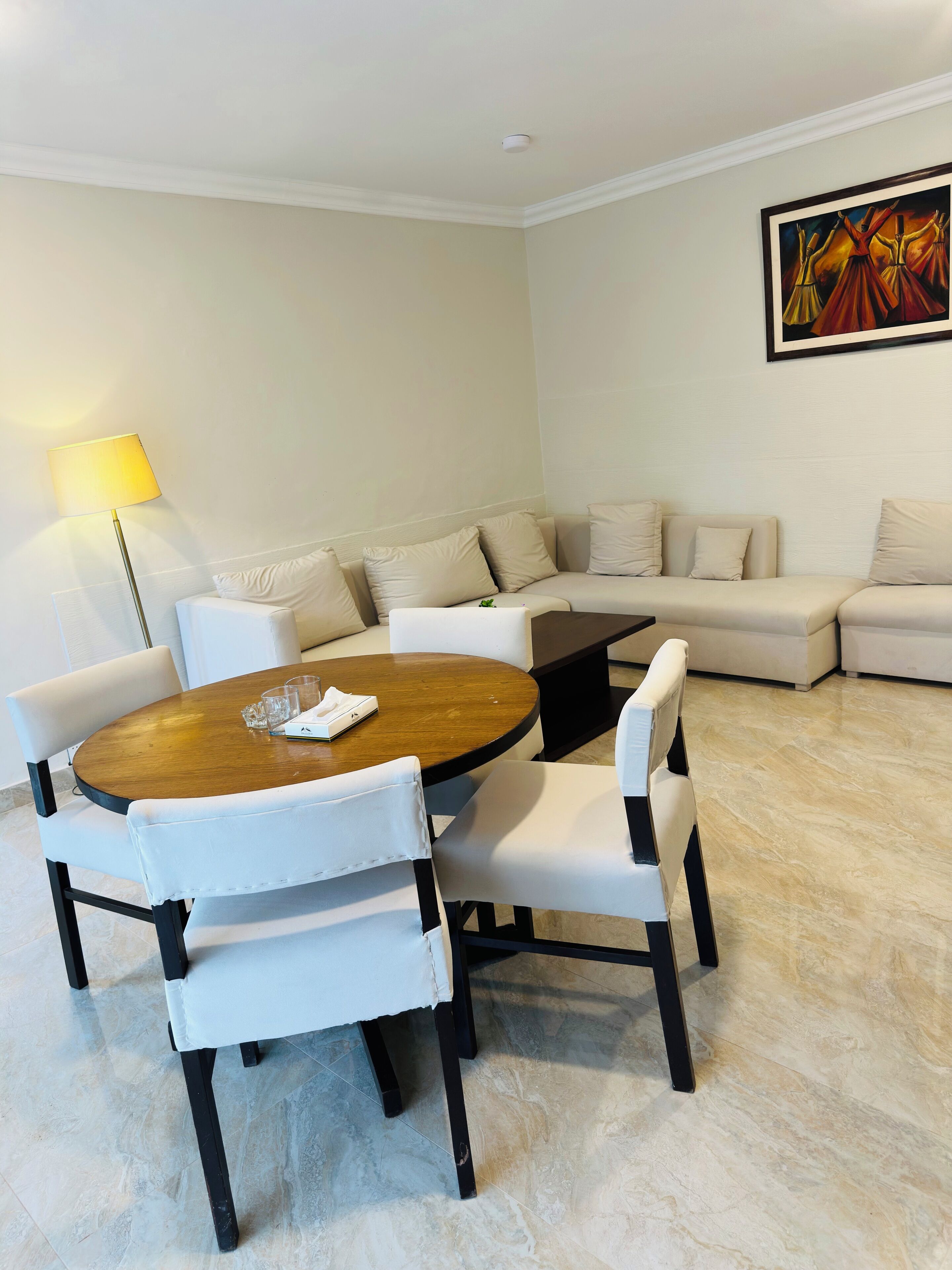 Uma sala de estar moderna com uma mesa redonda de madeira, cadeiras acolchoadas brancas, um sofá bege e um quadro na parede.