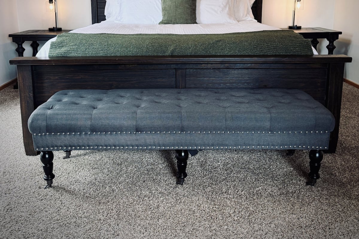 Quarto com teto de madeira, estrutura de cama de madeira escura, manta verde, duas luminárias de mesa e um banco cinzento.