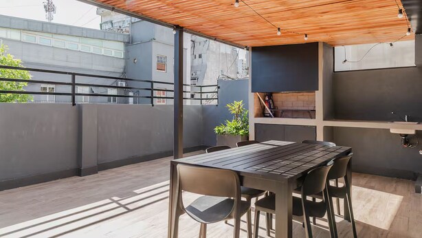 Um pátio externo com teto de madeira, mesa com cadeiras, churrasqueira e vista para edifícios.
