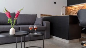 Uma sala de estar moderna com um sofá, uma mesa redonda pequena com um vaso e dois copos, e uma área de cozinha com bancada de mármore e armários pretos.