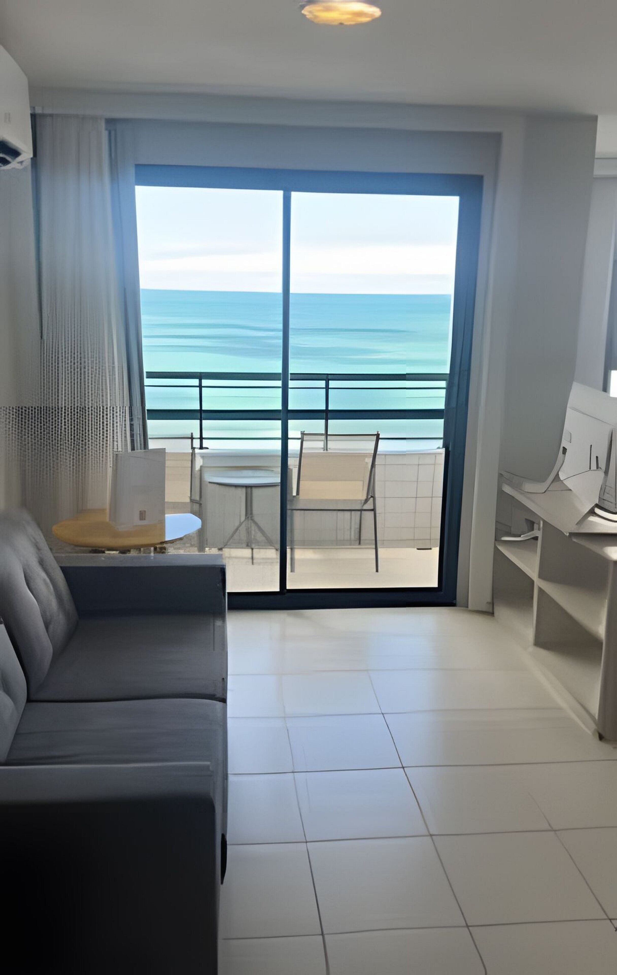 Quarto de hotel moderno com sofá cinzento, uma mesinha, uma cadeira e uma escrivaninha com computador. O quarto possui uma grande porta de vidro deslizante que dá para uma varanda com vista para o oceano.