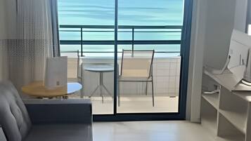 Quarto de hotel moderno com sofá cinzento, uma mesinha, uma cadeira e uma escrivaninha com computador. O quarto possui uma grande porta de vidro deslizante que dá para uma varanda com vista para o oceano.