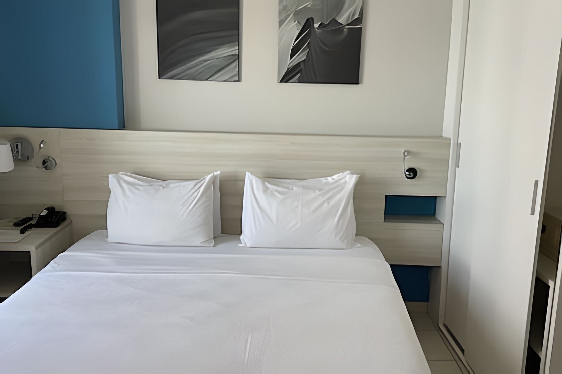 Quarto de hotel com cama, mesa de cabeceira, telefone e luminária fixada na parede.