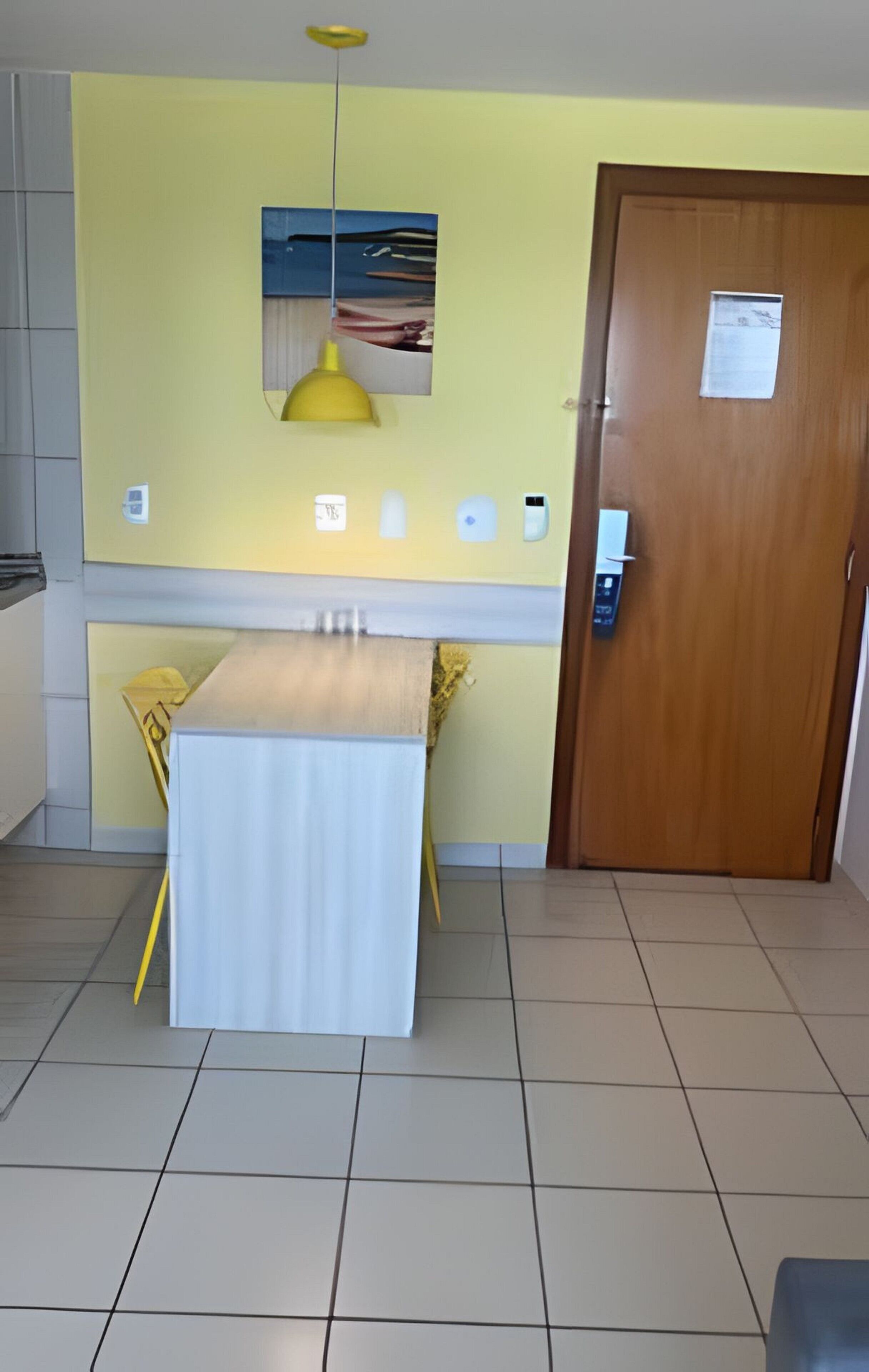 Um quarto com piso de azulejos, porta de madeira, mesa branca com cadeiras amarelas, luminária pendente amarela e uma pequena pintura na parede.