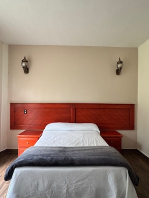 Quarto de hotel com cabeceira de madeira, duas luminárias fixadas na parede, cama com manta cinza e mesa de cabeceira de madeira.