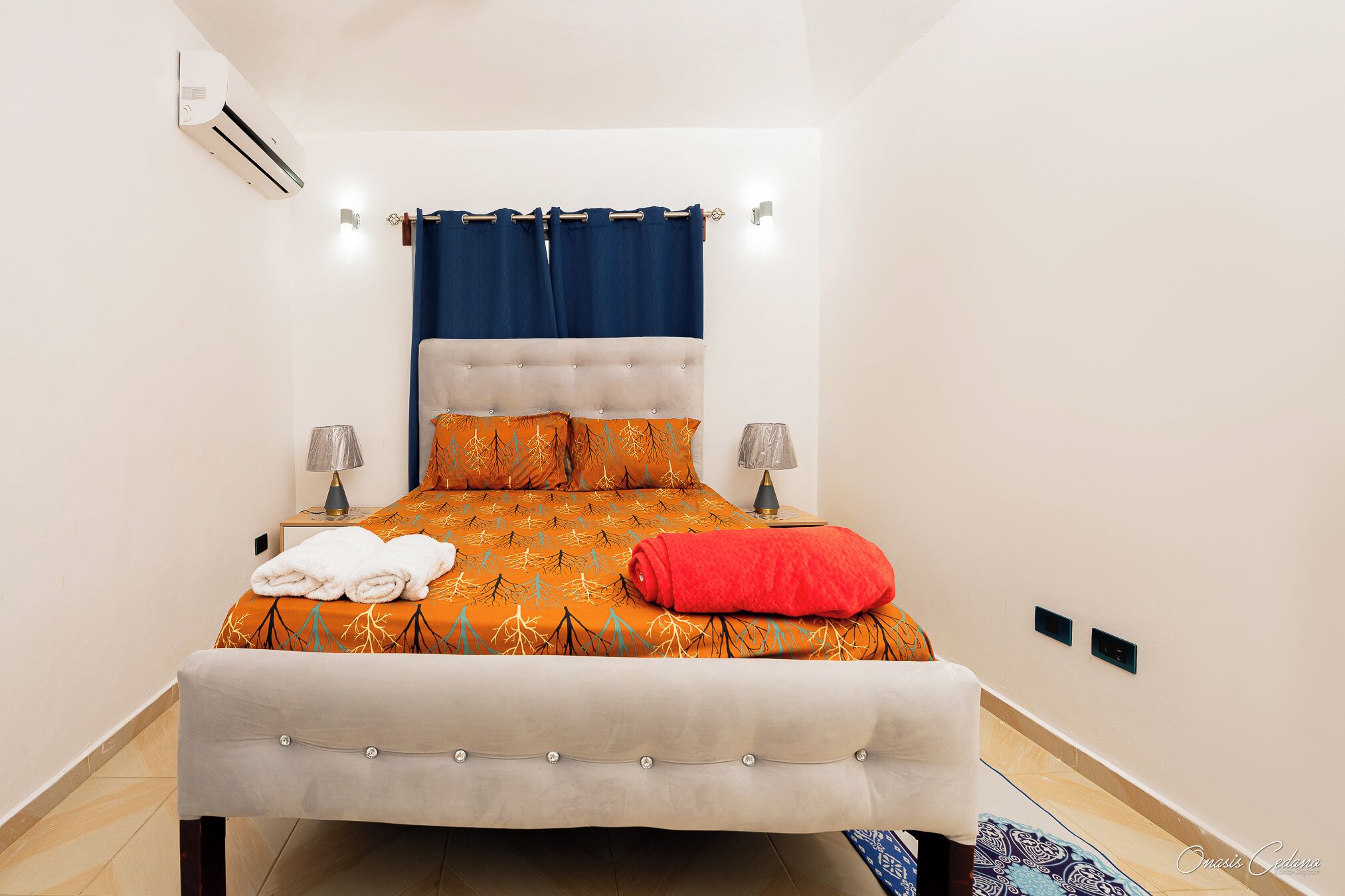 Uma cama bem arrumada com um edredom estampado, um travesseiro vermelho e duas luminárias de cabeceira.