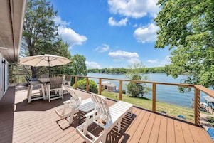 Um deck de madeira com móveis de exterior, uma mesa e cadeiras, com vista para um lago com árvores e um céu claro.