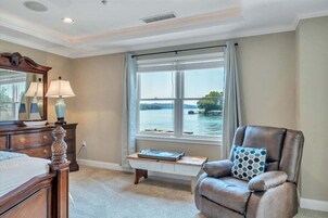 Quarto com uma janela grande, cama, cômoda, cadeira e mesa de cabeceira.